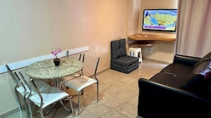 Um espaço de estar compacto com uma mesa de jantar redonda, duas cadeiras, um sofá preto, uma televisão e uma pequena orquídea em vaso.