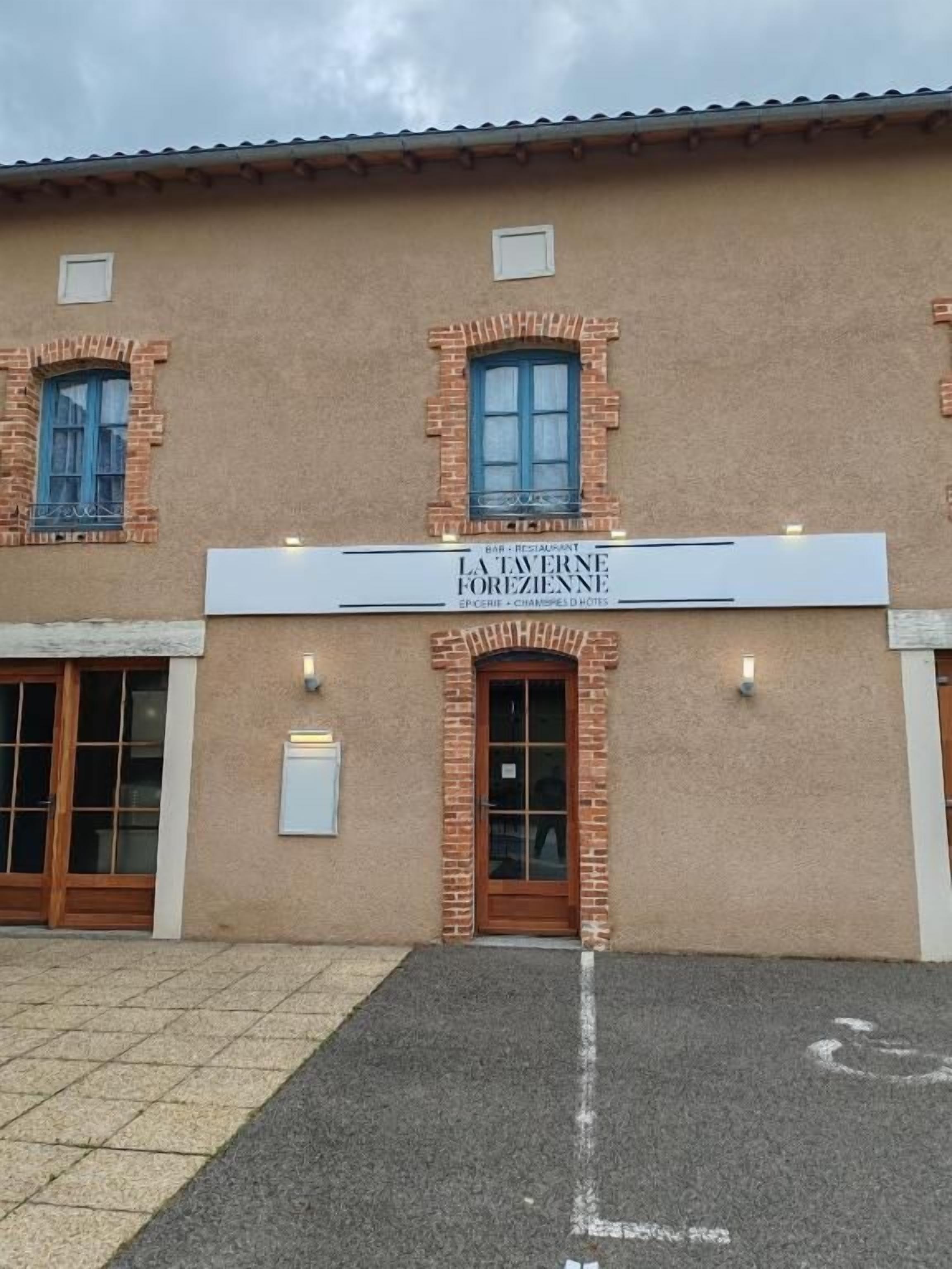 Um edifício com uma placa que diz 'LA TAVERNE FOREZIENNE', indicando um restaurante ou bar. Possui duas janelas grandes com molduras azuis, um arco de tijolos e uma porta.