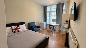 Quarto de hotel com cama, escrivaninha, cadeira, pequeno espaço de cozinha e televisão.