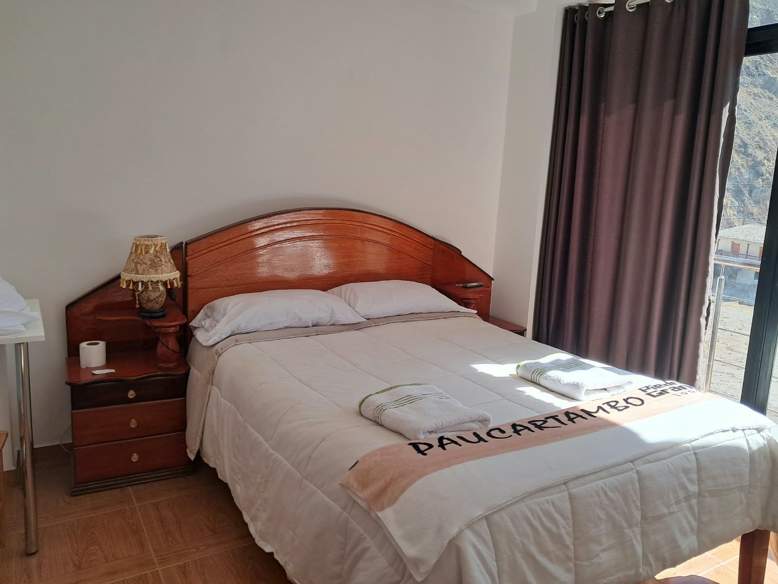 Quarto de hotel com cabeceira de madeira, cama com colcha branca, mesa de cabeceira com abajur e vista para o exterior através de uma janela com cortinas.