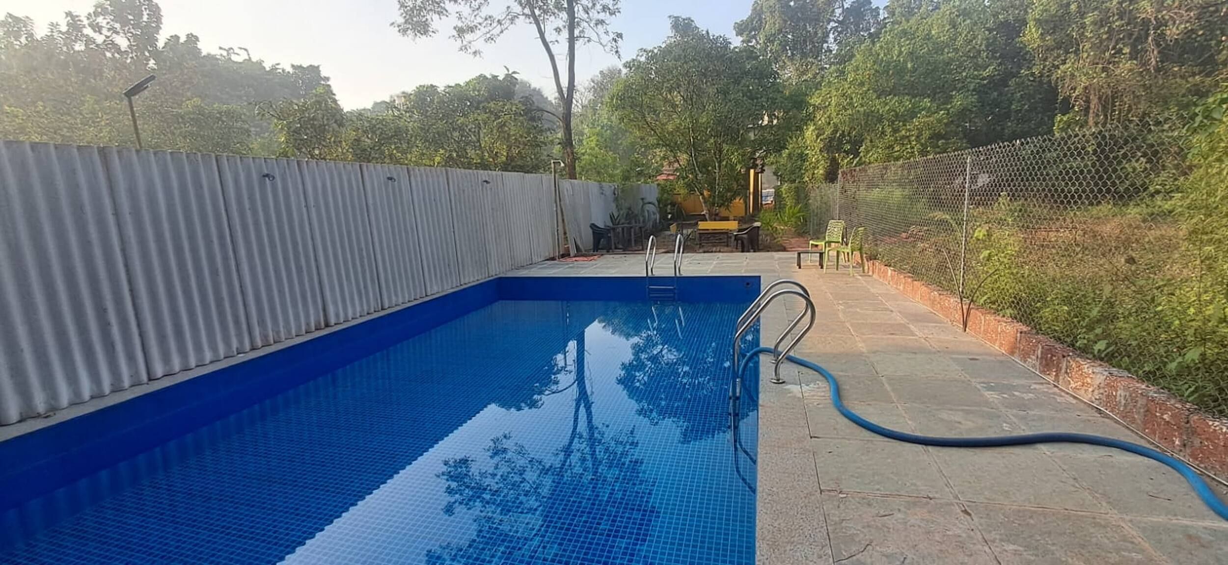 Piscina retangular com revestimento de azulejos azuis, cercada por uma grade de metal e um calçadão pavimentado.