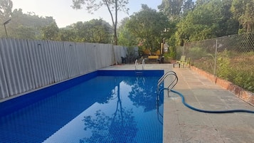 Piscina retangular com revestimento de azulejos azuis, cercada por uma grade de metal e um calçadão pavimentado.