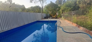 Piscina retangular com revestimento de azulejos azuis, cercada por uma grade de metal e um calçadão pavimentado.