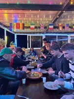 Um grupo de pessoas jantando à mesa, com vários pratos, bandeiras e vista para o céu noturno.