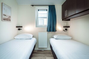 Quarto com duas camas de solteiro, janela com cortina, radiador e luminária na parede.