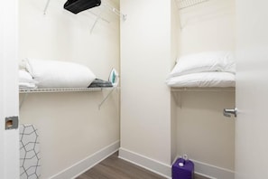 Quarto de hotel com cama, prateleira com roupas dobradas, cesto de roupa suja e tábua de passar roupa presa na parede.