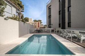 Piscina com cerca branca, espreguiçadeiras e um edifício ao fundo.