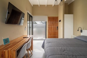 Quarto com cama, escrivaninha com cadeira, TV na parede e porta que dá para uma varanda.