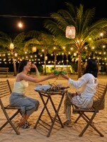 Duas pessoas sentadas à mesa em cenário tropical, com palmeiras e luzes de corda.