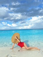 Uma pessoa sentada na praia, usando um chapéu grande, um maiô vermelho, com o oceano e o céu ao fundo.