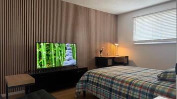 Quarto com televisão sobre um suporte, cama com colcha xadrez, mesa de cabeceira e janela com persianas.