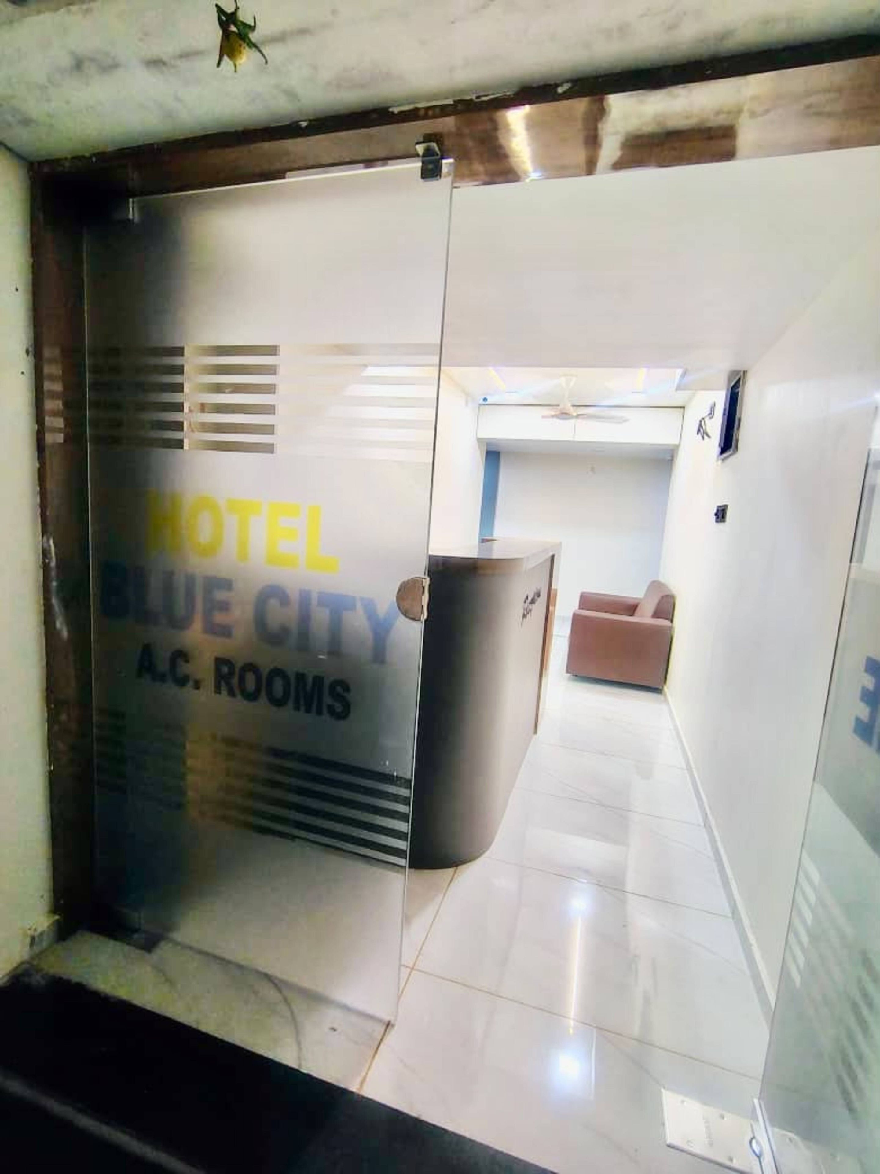 Entrada de hotel com balcão de recepção, área de estar e placa indicando 'HOTEL BLUE CITY A.C. ROOMS'.