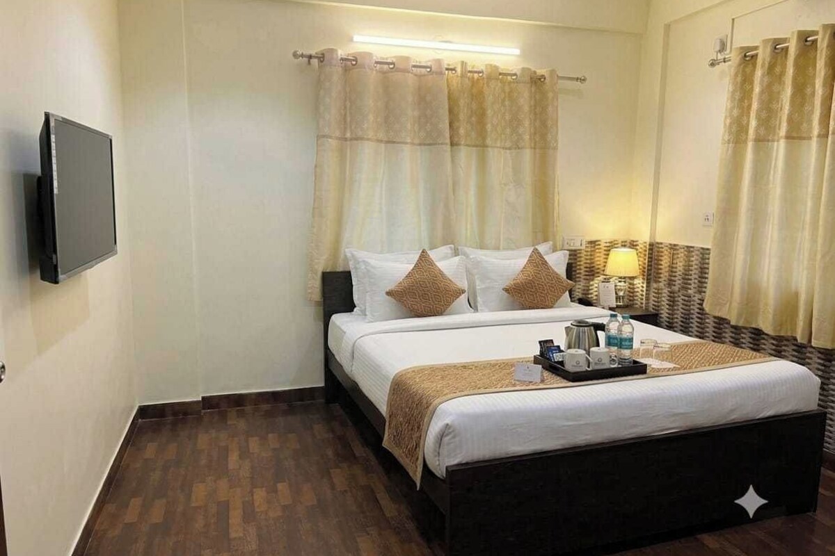 Quarto de hotel com cama, televisão de tela plana, mesa de cabeceira com luminária e janela com cortinas.