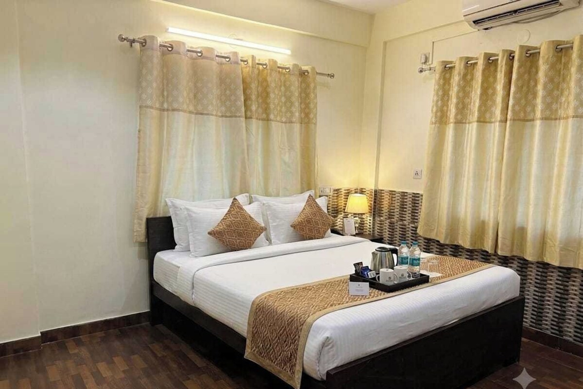 Quarto de hotel com cama, cortinas, mesa de cabeceira com abajur e bandeja com artigos de higiene.