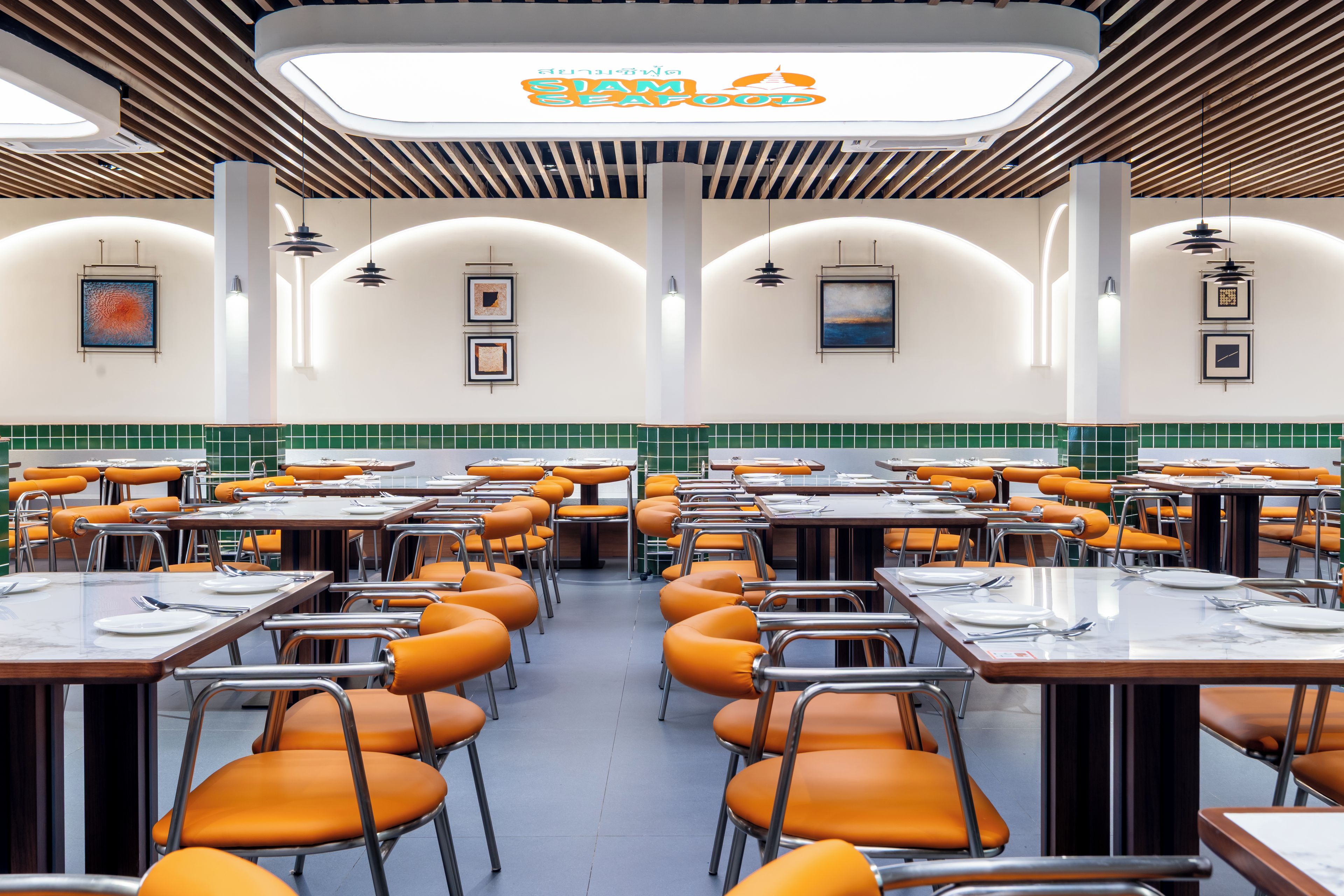 Interior de restaurante com várias mesas e cadeiras, painel de azulejos e iluminação pendente.