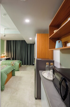 Quarto de hotel compacto com cozinha compacta, cama com roupa de cama verde, escrivaninha e uma grande janela com cortinas.