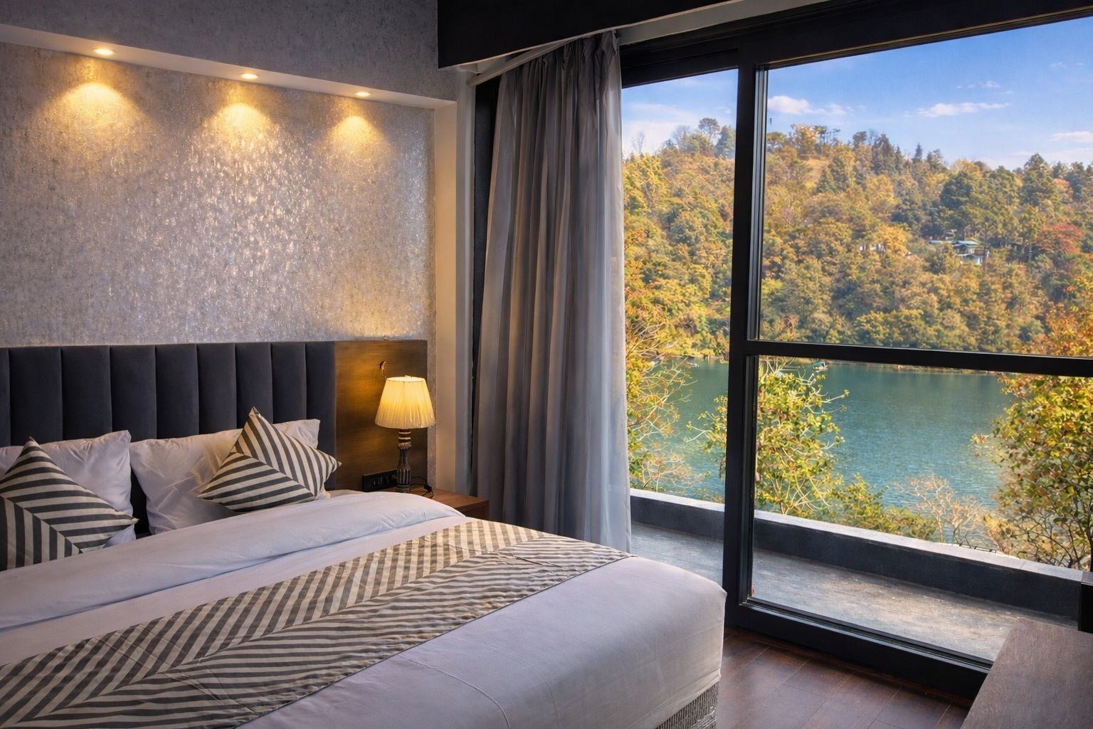 Quarto com cama grande, mesa de cabeceira, abajur e vista para um lago rodeado de árvores.