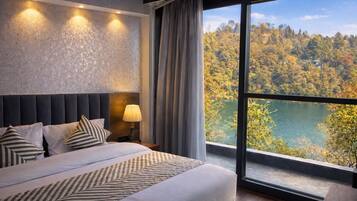 Quarto com cama grande, mesa de cabeceira, abajur e vista para um lago rodeado de árvores.