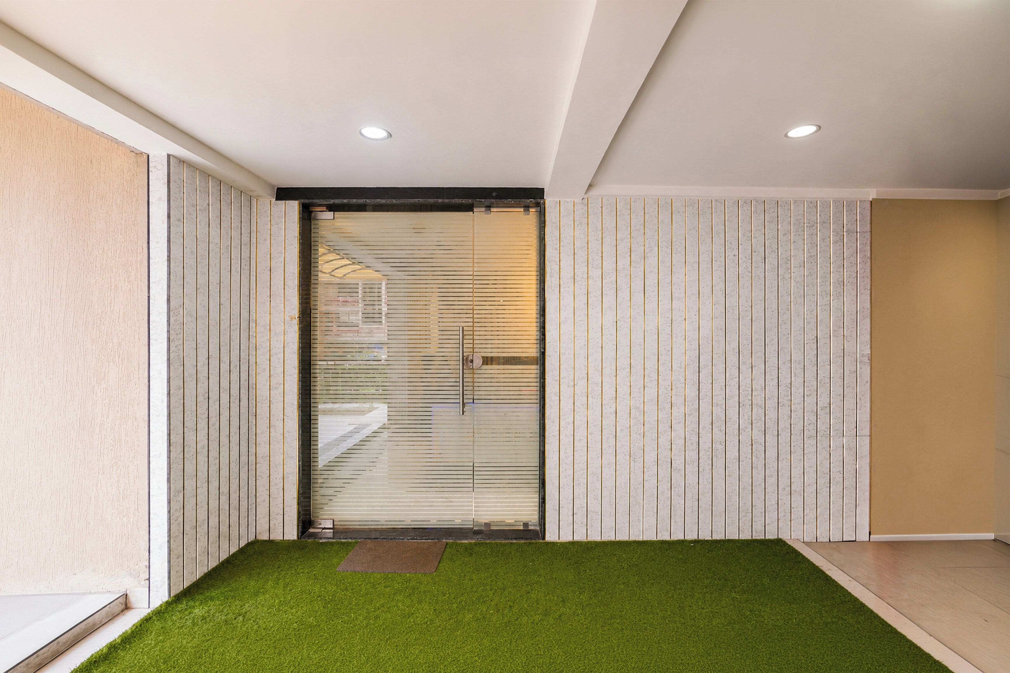 Interior moderno com entrada por porta de vidro, área com grama sintética verde e parede bege com linhas verticais.