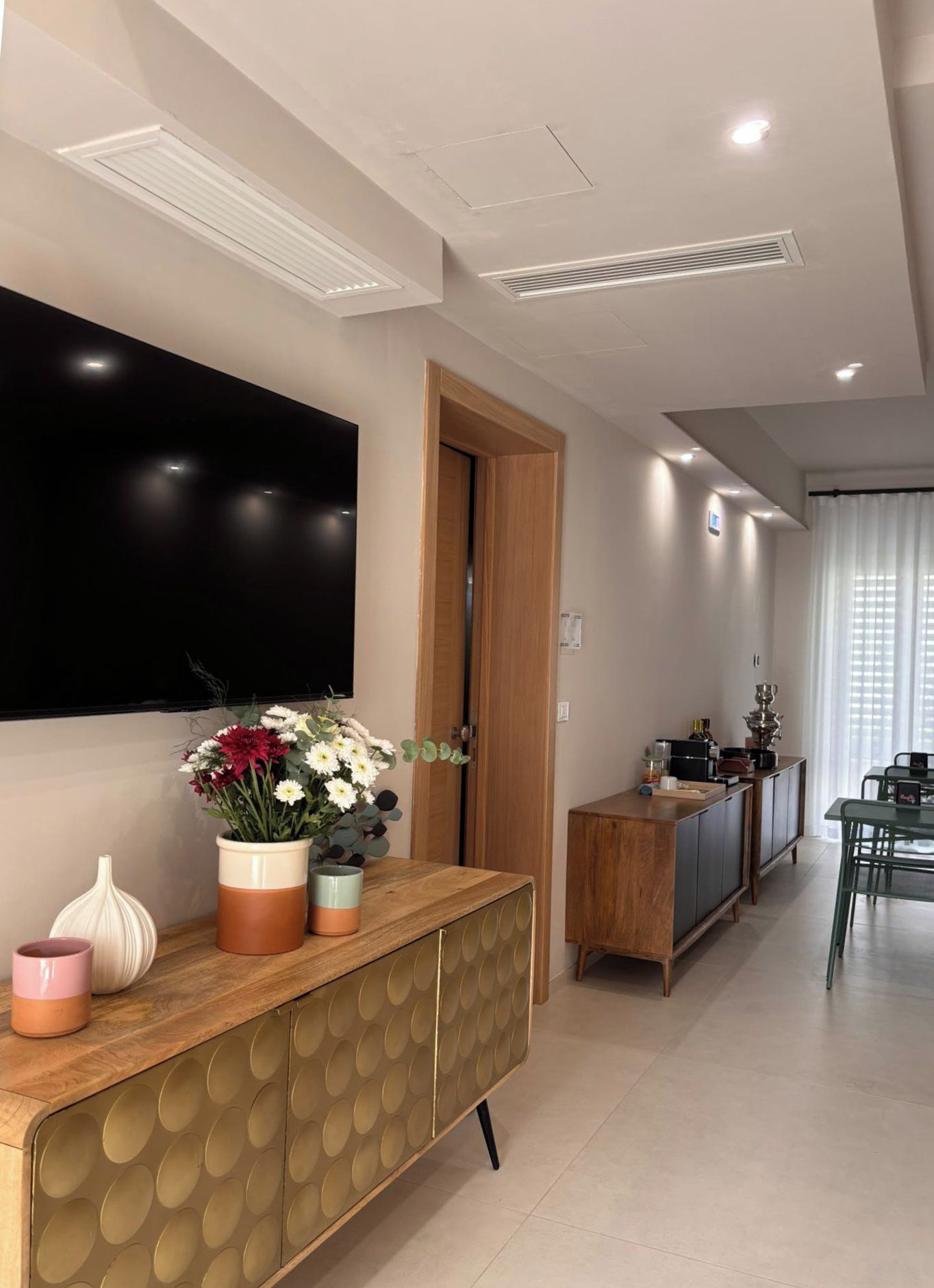 Uma sala de estar moderna com televisão de tela plana na parede, uma mesa consola de madeira com objetos decorativos, uma área de jantar com mesa e cadeiras, e uma cozinha com armários e bancada.
