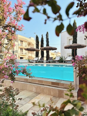Piscina com espreguiçadeiras e guarda-sóis, cercada por edifícios e vegetação.