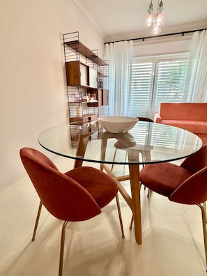 Uma área de refeições com mesa de vidro, cadeiras vermelhas, estante de madeira e janela com persianas.