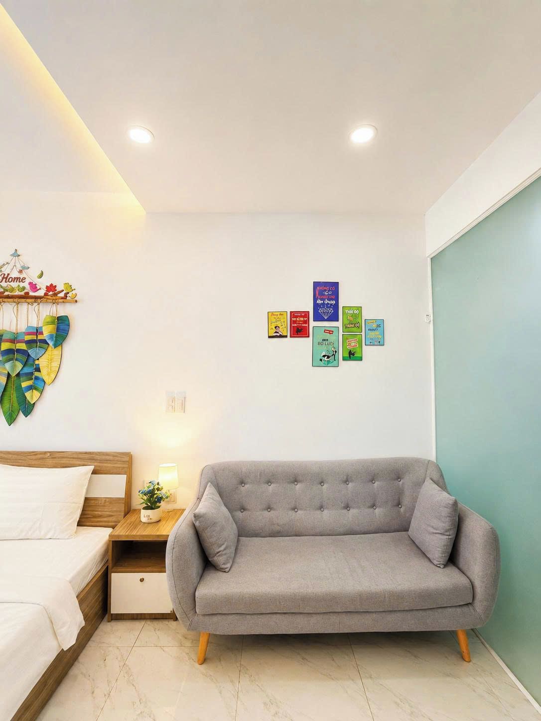 Quarto com cama, sofá cinza, mesa de cabeceira, parede com quadros e luminária no teto.