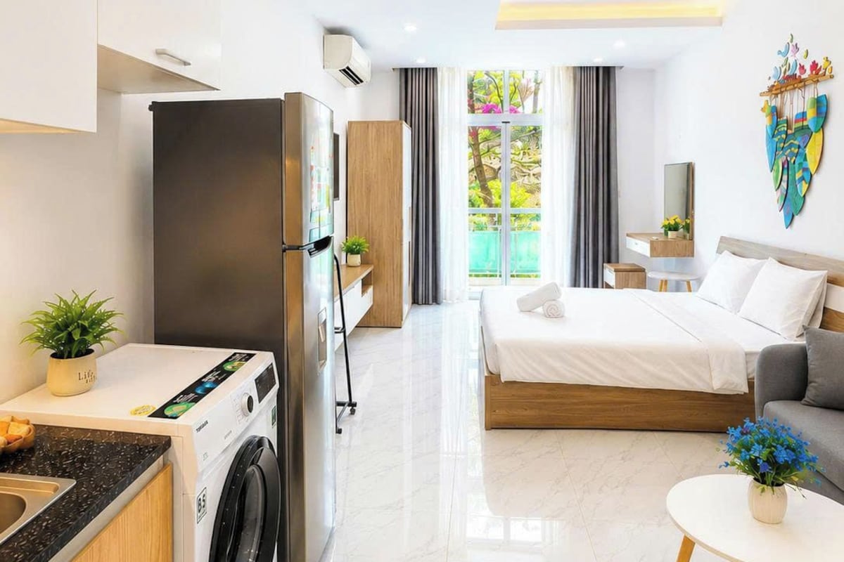 Quarto de hotel moderno com cama, sofá, mesa de jantar, máquina de lavar e área de cozinha com geladeira.