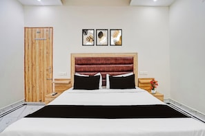Quarto de hotel com porta de madeira, cama com cabeceira vermelha, mesa de cabeceira com um vaso de flores e três obras de arte emolduradas na parede.