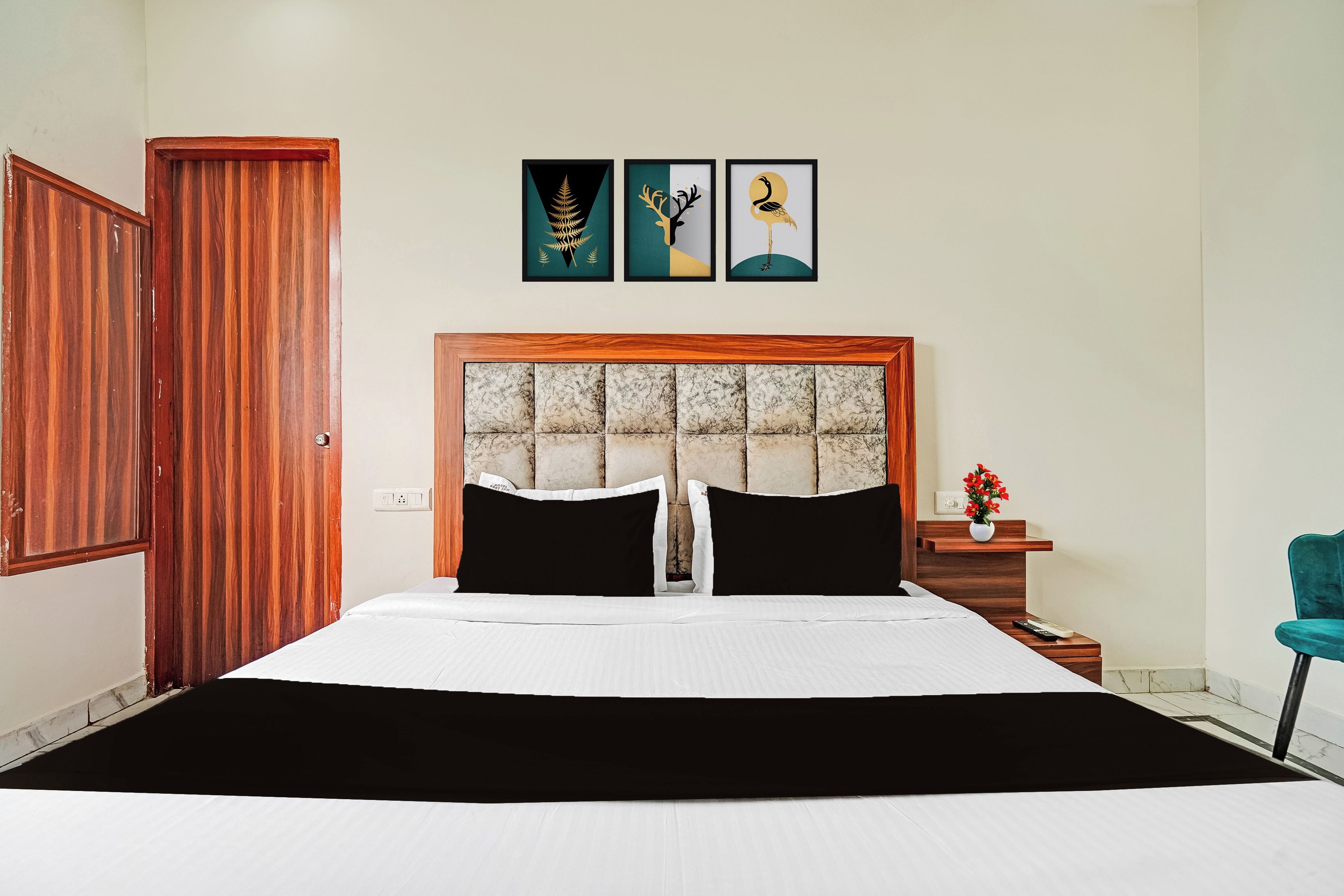 Quarto com cabeceira de madeira, cama com roupa de cama preta e branca, mesa de cabeceira com um vaso de flores e três obras de arte emolduradas na parede.