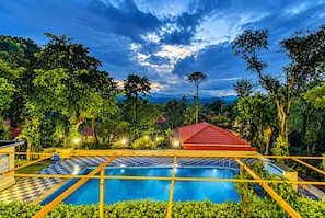 Piscina com deck quadriculado, cercada por vegetação exuberante e um edifício com telhado vermelho.