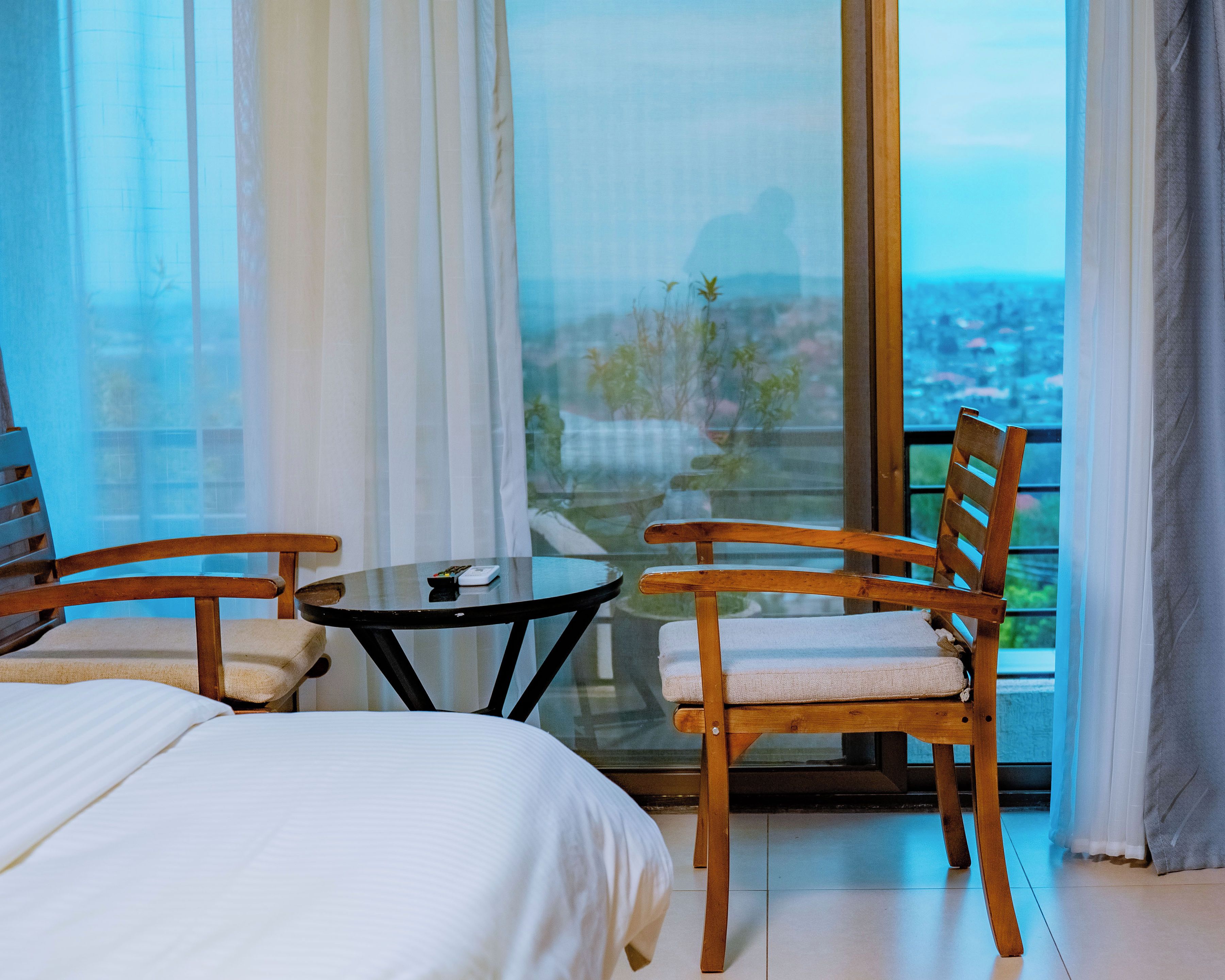 Quarto de hotel com varanda, uma cama, duas cadeiras de madeira, uma mesinha e vista para a paisagem urbana.