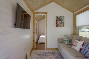 Quarto aconchegante com teto de madeira, uma cama, um sofá com almofadas estampadas e uma pintura de uma praia.