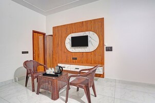 Quarto de hotel com parede de painel de madeira, televisão de tela plana fixada em superfície que imita mármore, mesa pequena com chaleira, duas cadeiras de vime e piso de azulejos brancos.