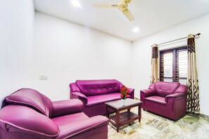 Quarto com ventoinha no teto, janela com cortinas estampadas e móveis roxos.