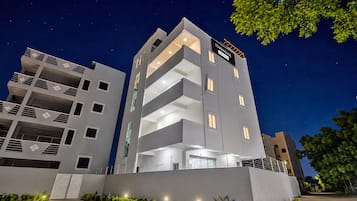 Um edifício de vários andares com varandas, uma placa indicando que é uma casa geminada com serviços, e uma entrada bem iluminada.