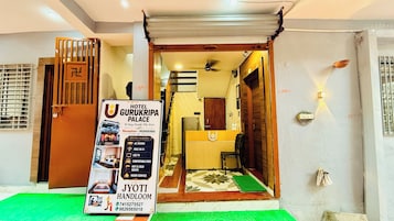 Entrada de hotel com balcão de recepção, placa indicando comodidades como quartos com ar condicionado e Wi-Fi, e um tapete verde na entrada.