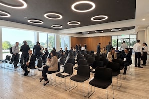 Sala de conferências com design moderno, parede revestida de madeira, teto alto com iluminação circular e uma grande janela com vista para o exterior.