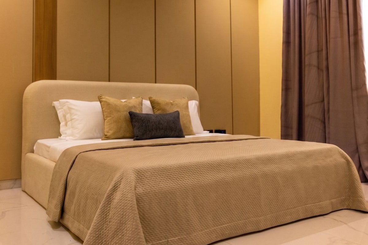 Cama com cabeceira bege e travesseiros, cobertor marrom e parede de painéis de madeira atrás.