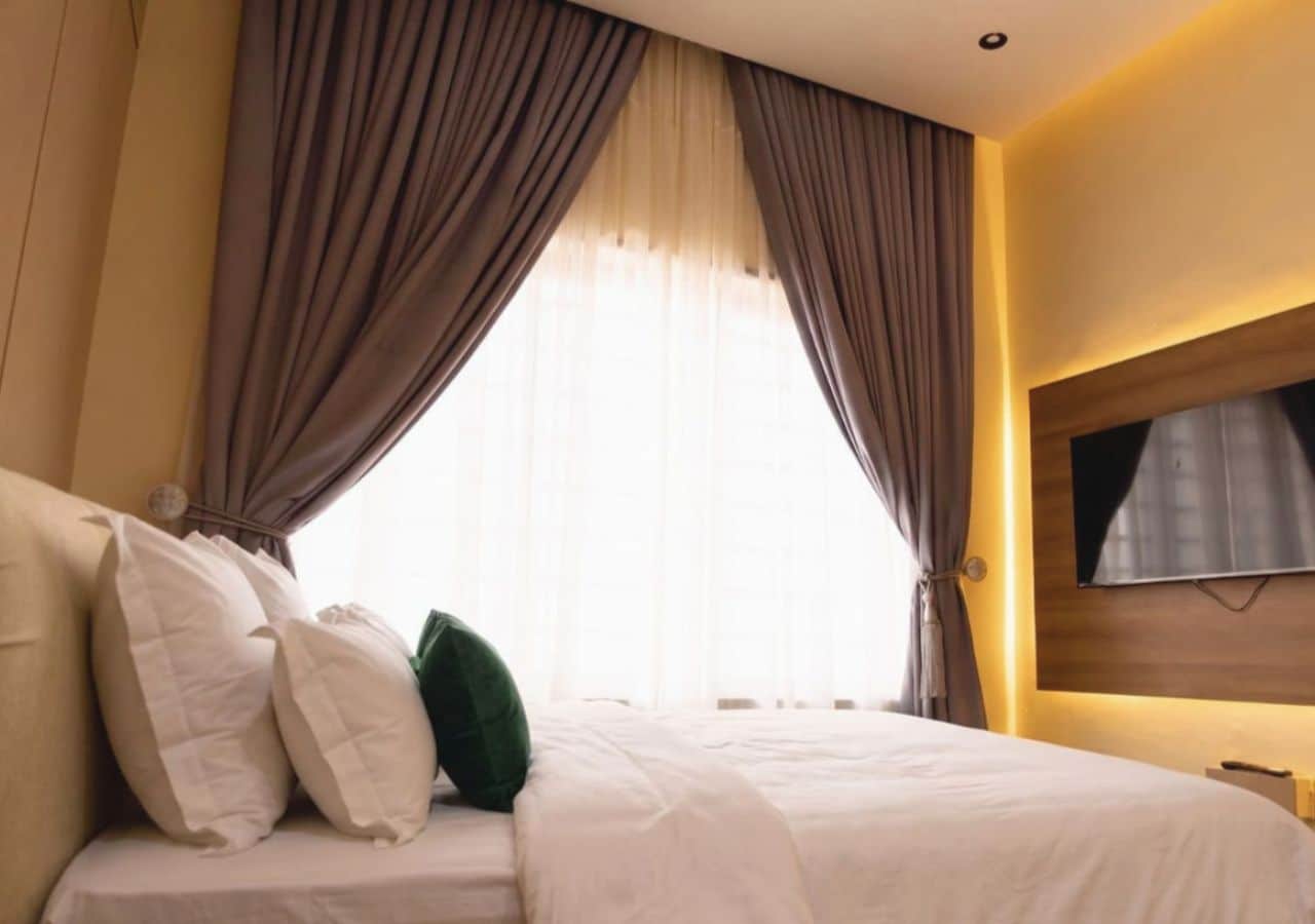 Quarto de hotel com cama, cortinas, televisão na parede e janela com cortinas leves.