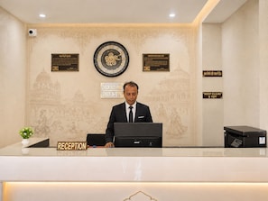 Bancada de recepção com um homem atrás, um relógio e uma parede com obras de arte e placas culturais.
