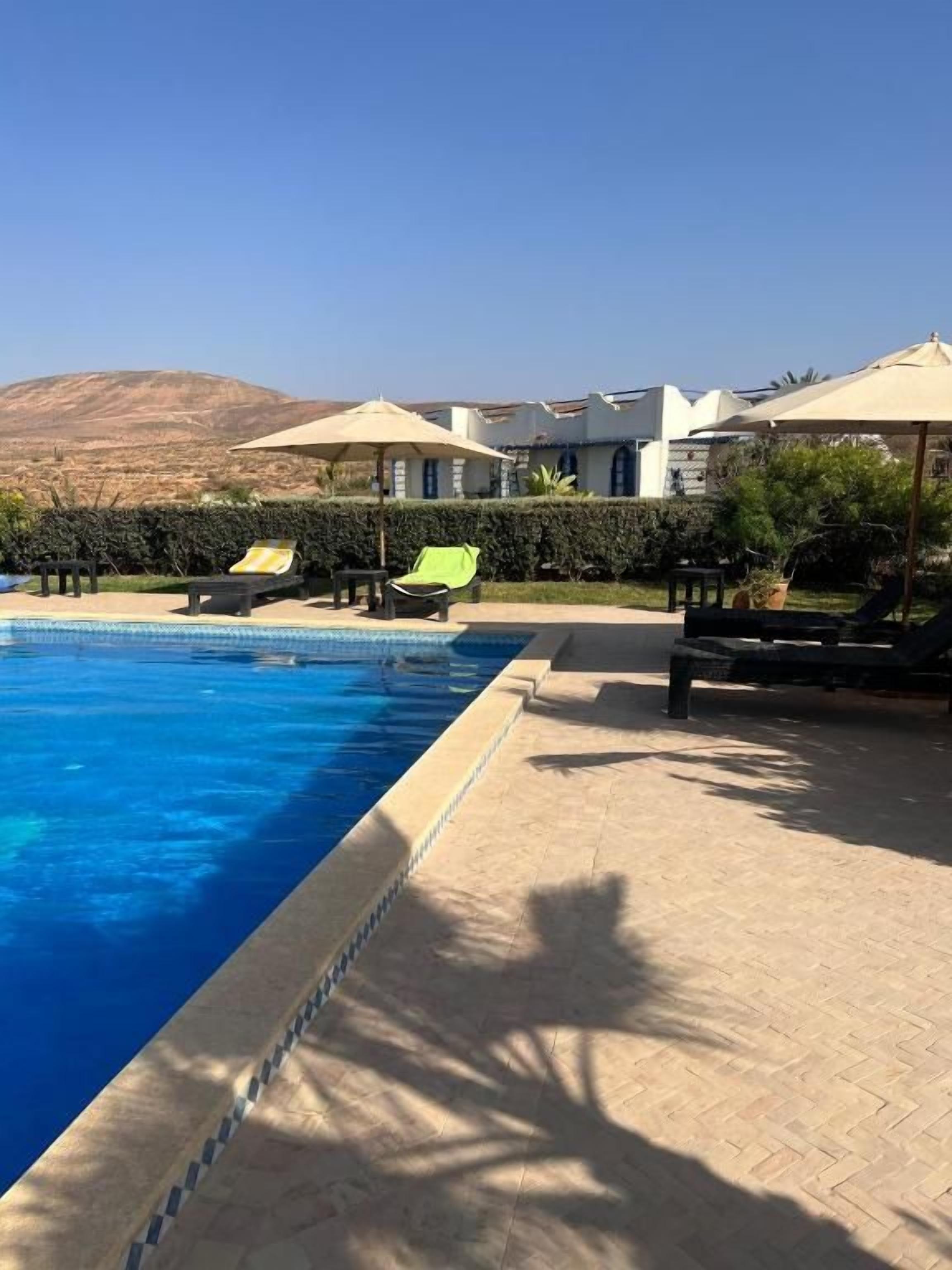 Piscina com espreguiçadeiras e guarda-sóis, em área ajardinada com vista para colinas ao longe.