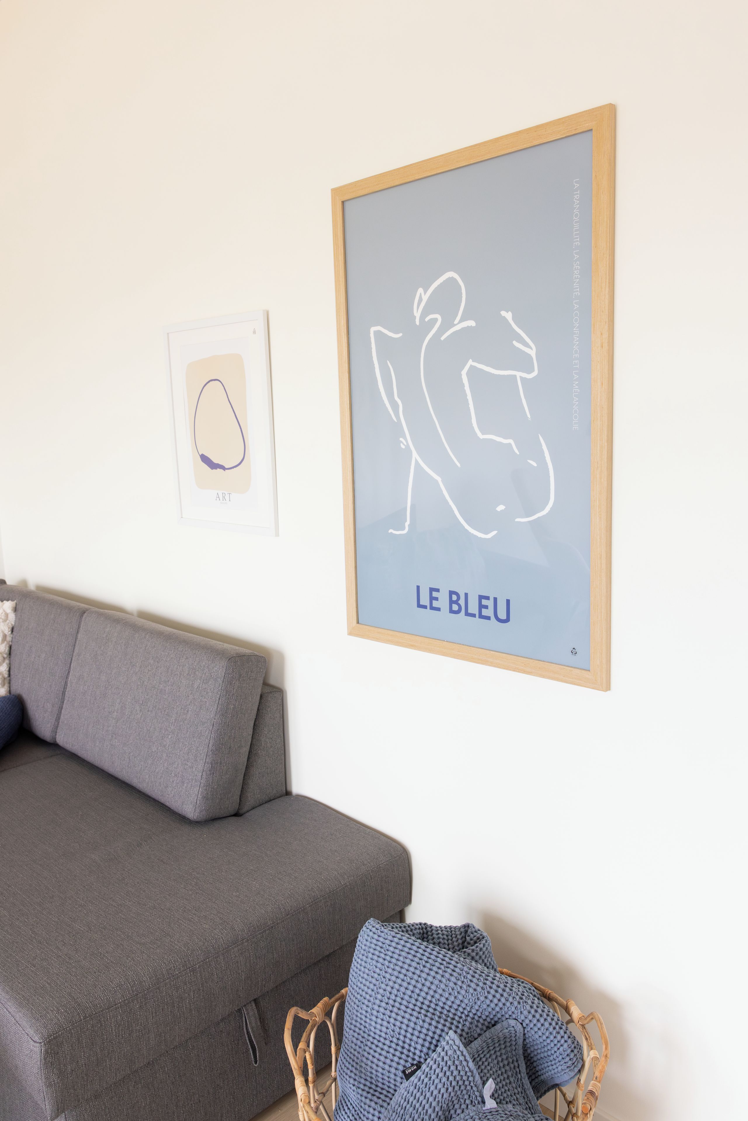 Um quarto com um sofá cinzento, uma obra de arte emoldurada com o texto 'LE BLEU' e uma peça pequena emoldurada com um design abstrato.
