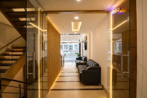 Um moderno hall de hotel com divisória de vidro, escada, área de estar com sofás e vista para a rua.