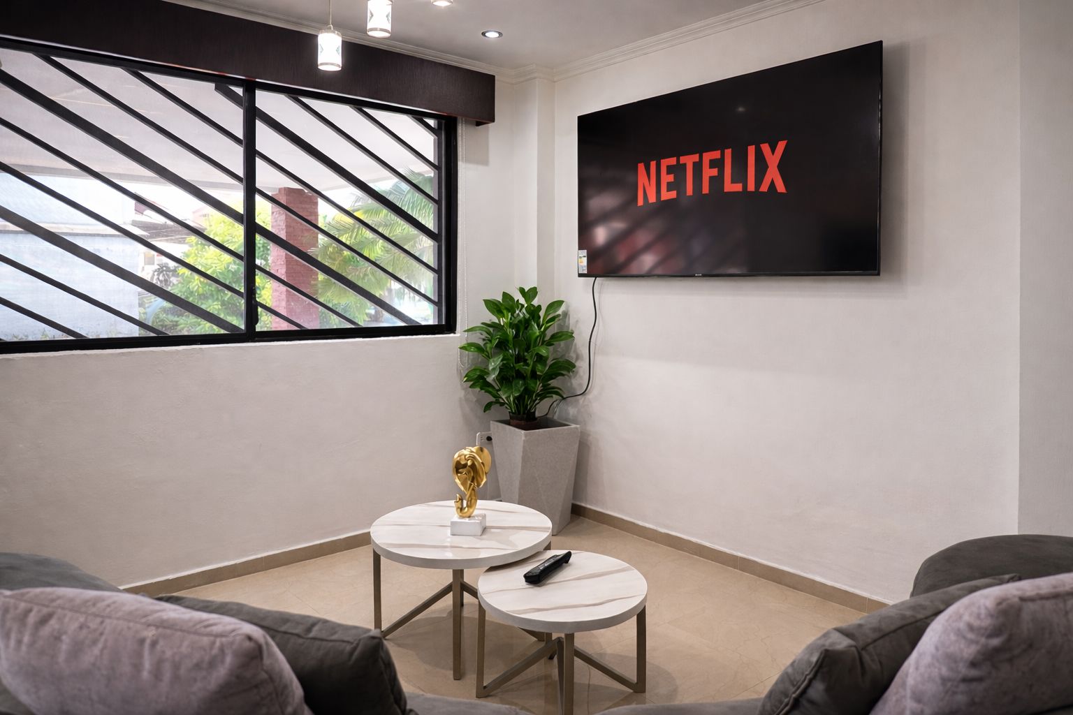 Quarto moderno com uma janela grande, TV fixada na parede exibindo 'NETFLIX', uma mesa redonda pequena com um controle remoto, uma planta em vaso e sofás cinzas.