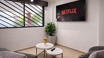 Quarto moderno com uma janela grande, TV fixada na parede exibindo 'NETFLIX', uma mesa redonda pequena com um controle remoto, uma planta em vaso e sofás cinzas.