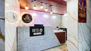 Uma área de recepção de hotel com parede de mármore, balcão de recepção e teto com vigas aparentes e luminárias pendentes.