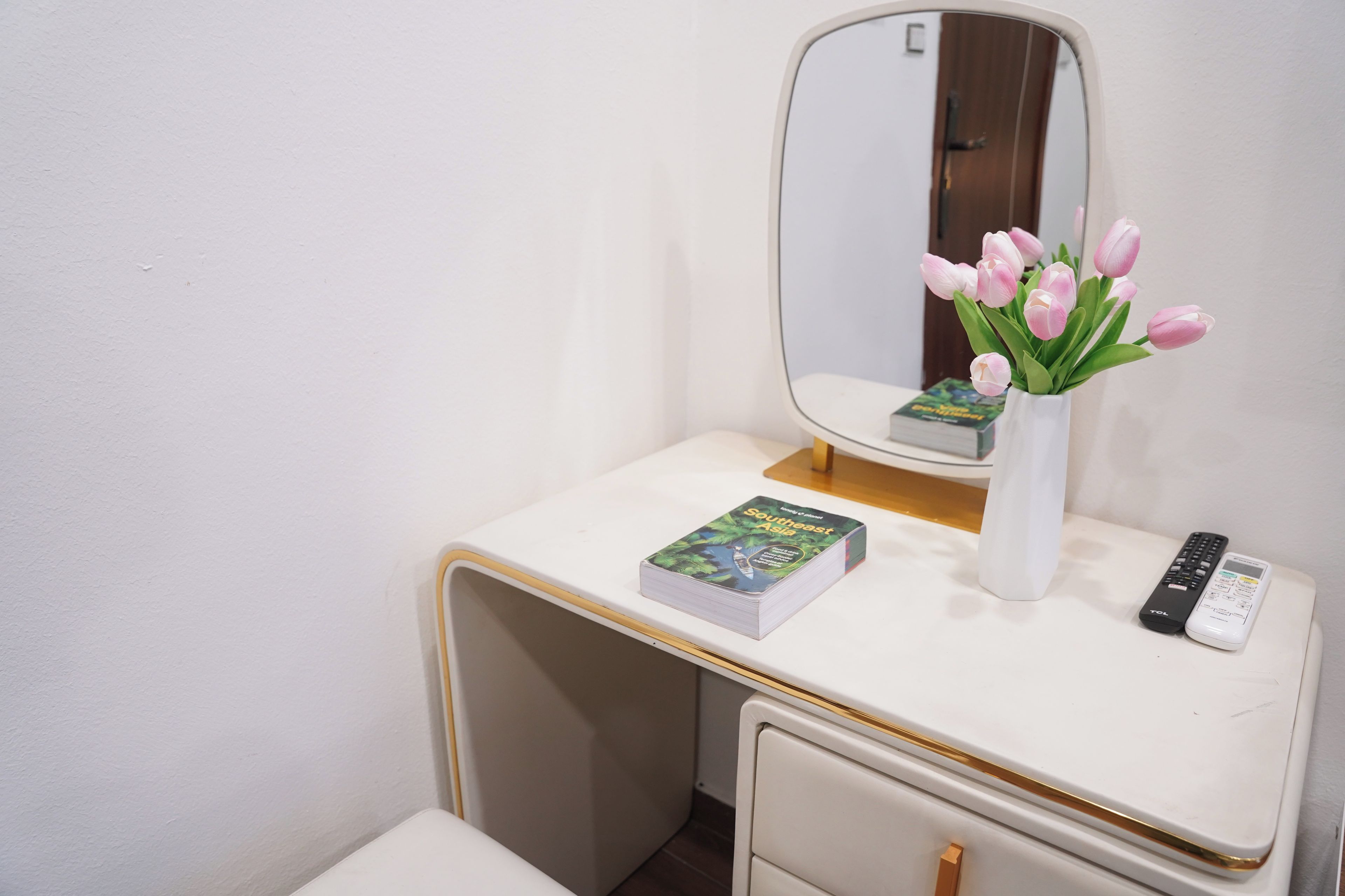 Uma toucador branca com espelho, um livro intitulado 'Sudeste Asiático', um vaso com flores cor-de-rosa e um controle remoto.