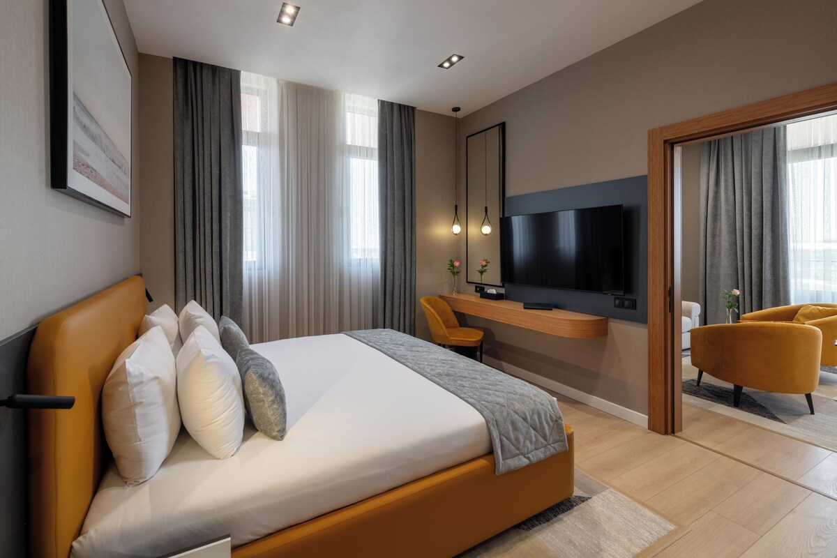 Quarto de hotel moderno com uma cama, uma escrivaninha com televisão de tela plana, uma cadeira e uma janela grande com cortinas.
