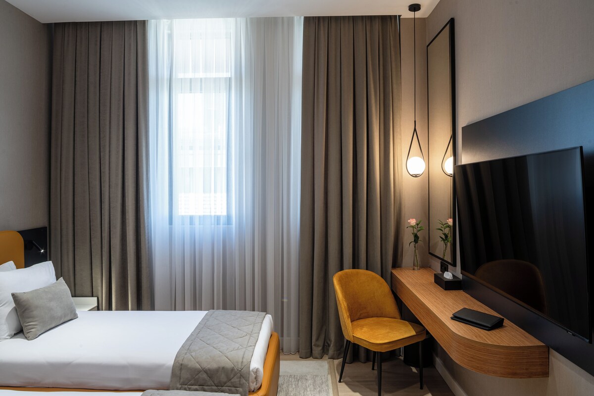 Quarto de hotel com duas camas, uma escrivaninha, uma cadeira, uma TV e uma janela grande com cortinas.